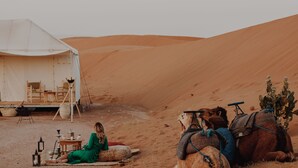 Tenda no deserto com mesa e cadeiras montadas para uma refeição, rodeada por dunas de areia.