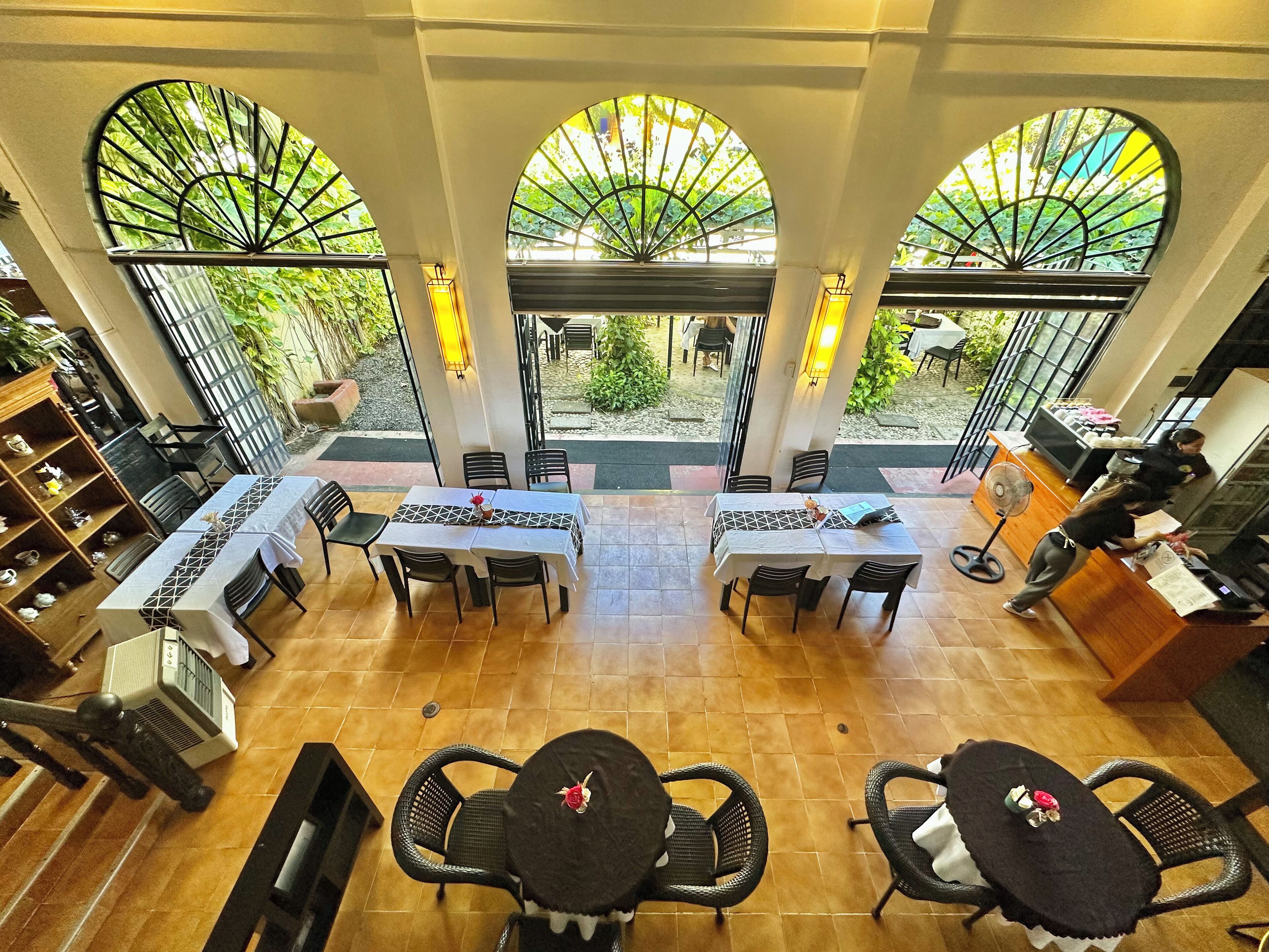 Restaurante com mesas e cadeiras dispostas para refeições, amplas janelas em arco com vegetação lá fora e um balcão com cardápio.