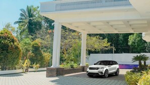 Uma entrada coberta com um carro branco estacionado, rodeada por vegetação e um edifício branco com telhado vermelho.