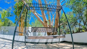 Um banco com estrutura metálica e ripas de madeira, localizado numa área pavimentada com uma árvore ao fundo.
