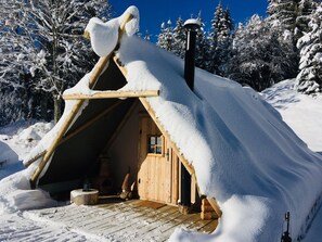 Uma cabana de madeira coberta de neve, com telhado inclinado, porta de madeira e um pequeno alpendre.