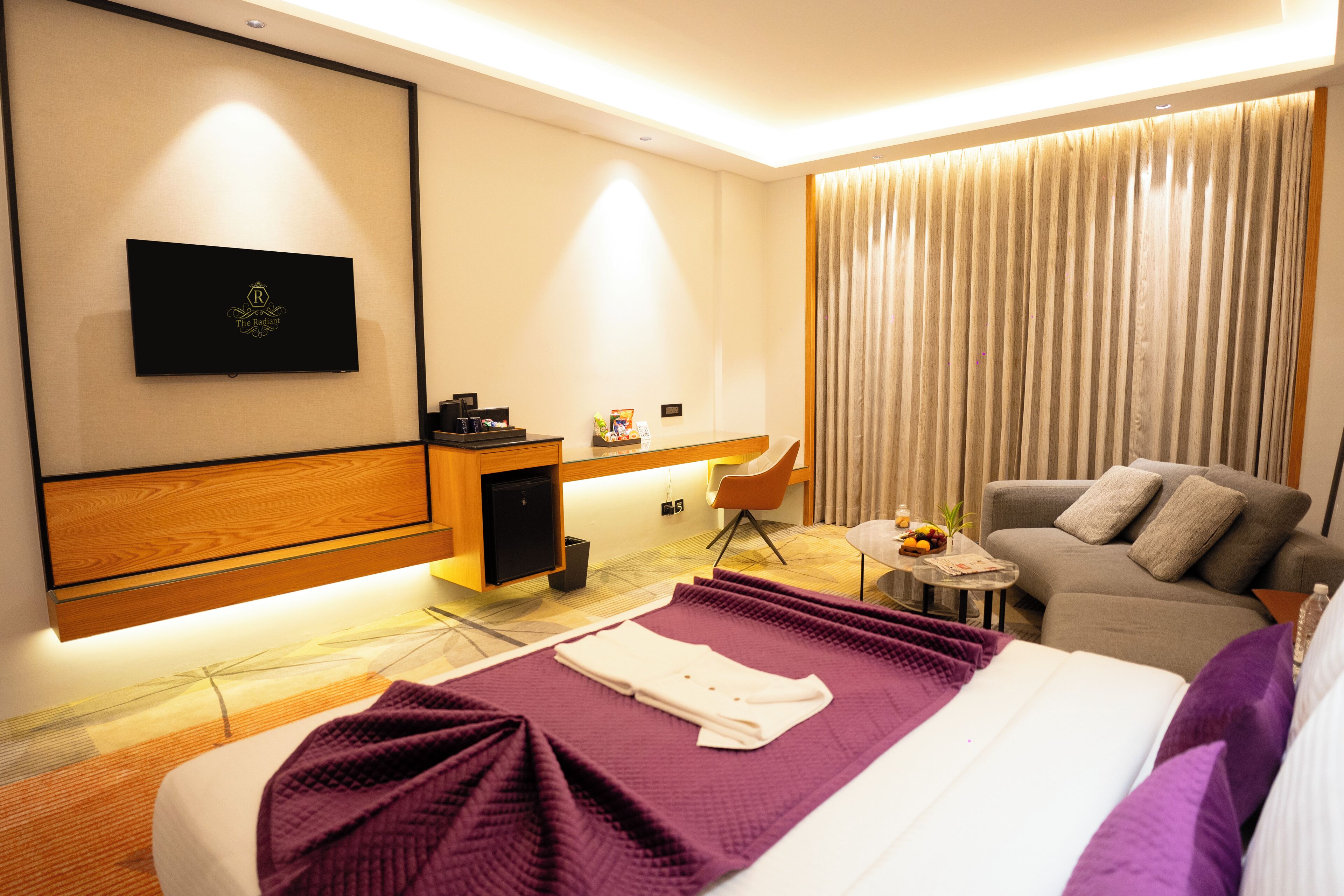 Quarto de hotel moderno com uma cama bem arrumada, uma escrivaninha com computador, uma poltrona confortável e uma janela grande com cortinas.