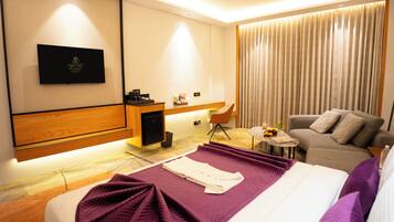 Quarto de hotel moderno com uma cama bem arrumada, uma escrivaninha com computador, uma poltrona confortável e uma janela grande com cortinas.