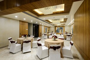 Uma sala de banquetes com mesas redondas, cadeiras brancas com fitas castanhas e lustres.