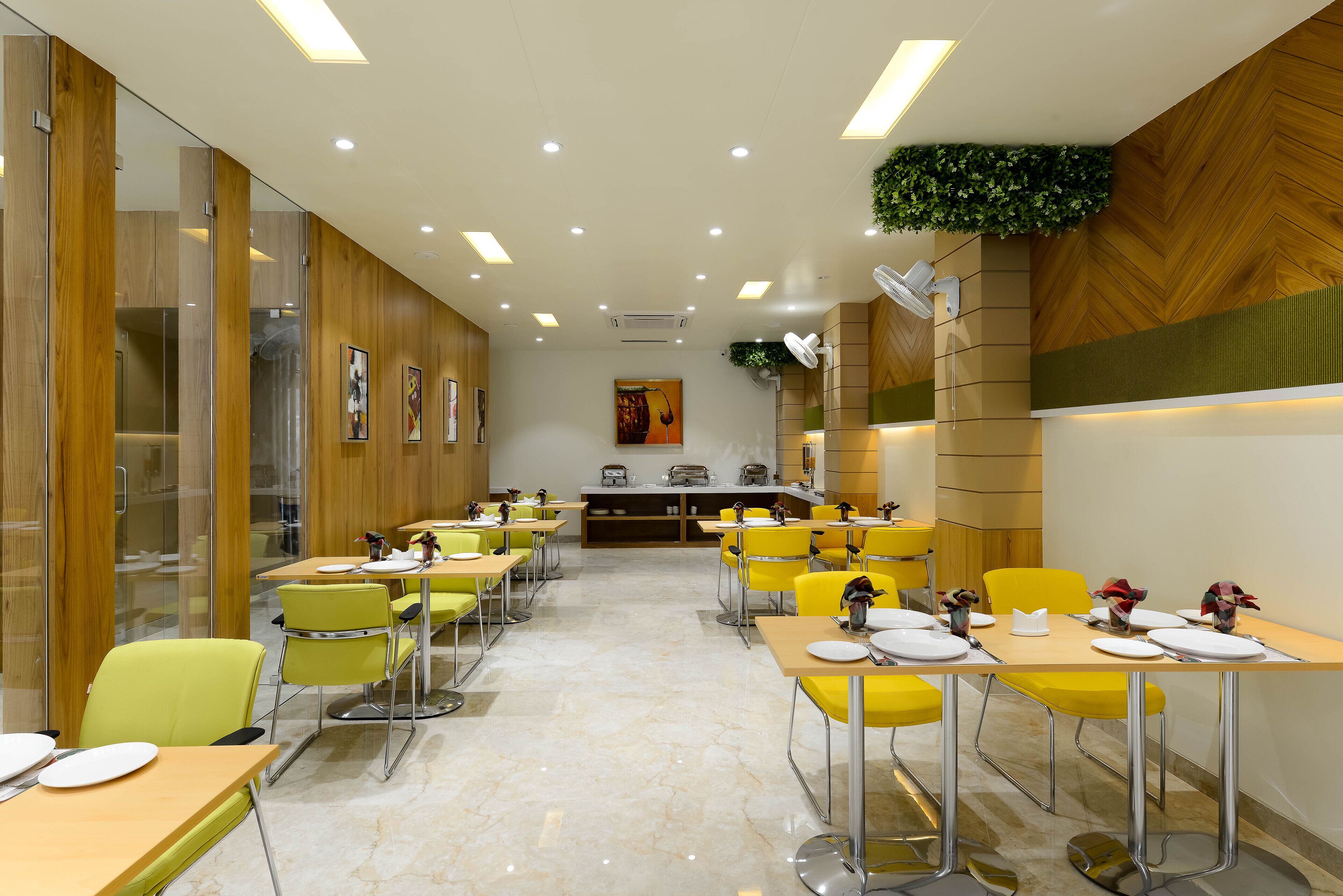 Uma área de refeições moderna com cadeiras e mesas amarelas, uma cozinha ao fundo e uma parede de madeira com uma planta verde.