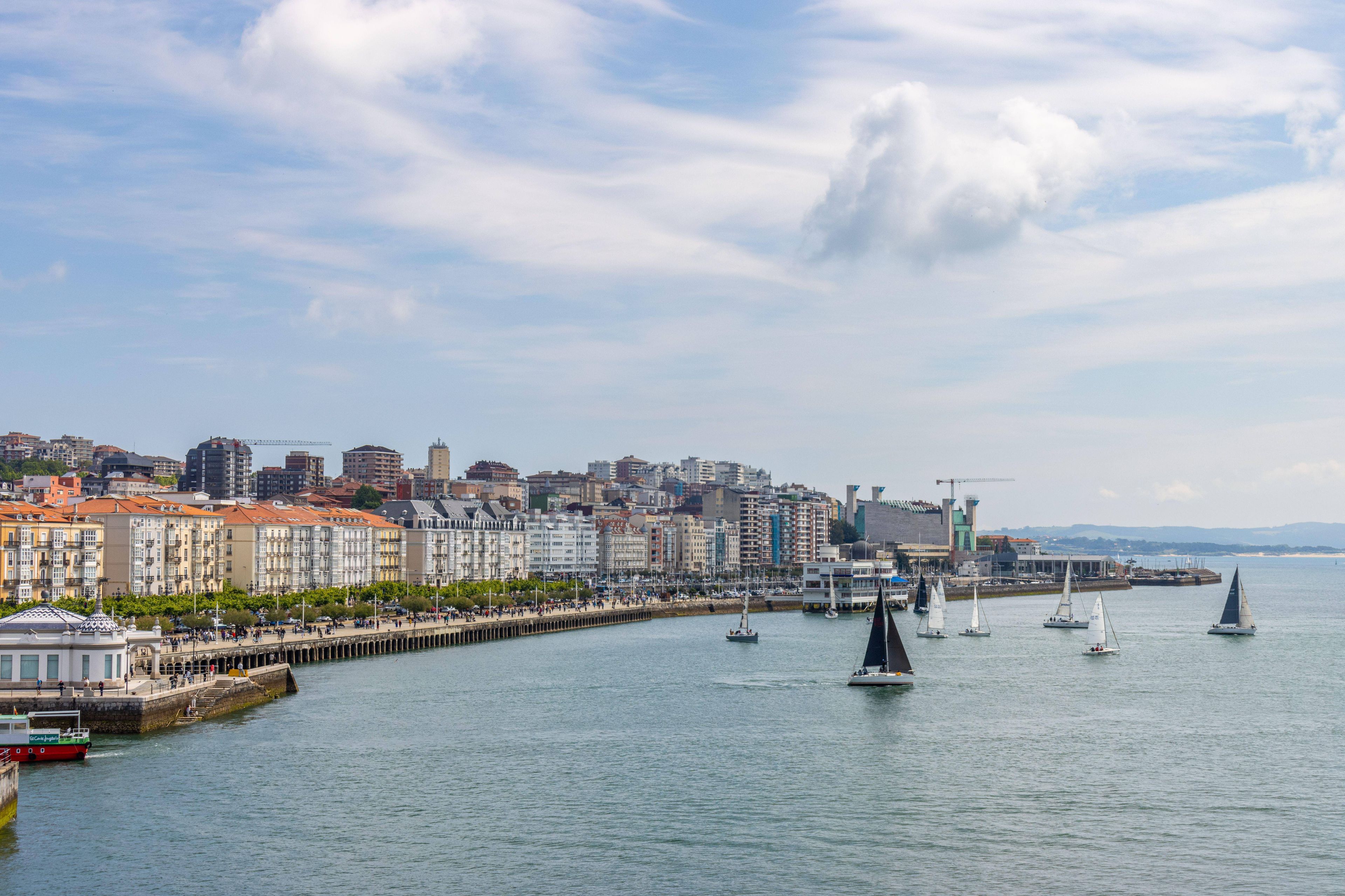 Paisagem urbana à beira-mar com edifícios, um cais e veleiros.