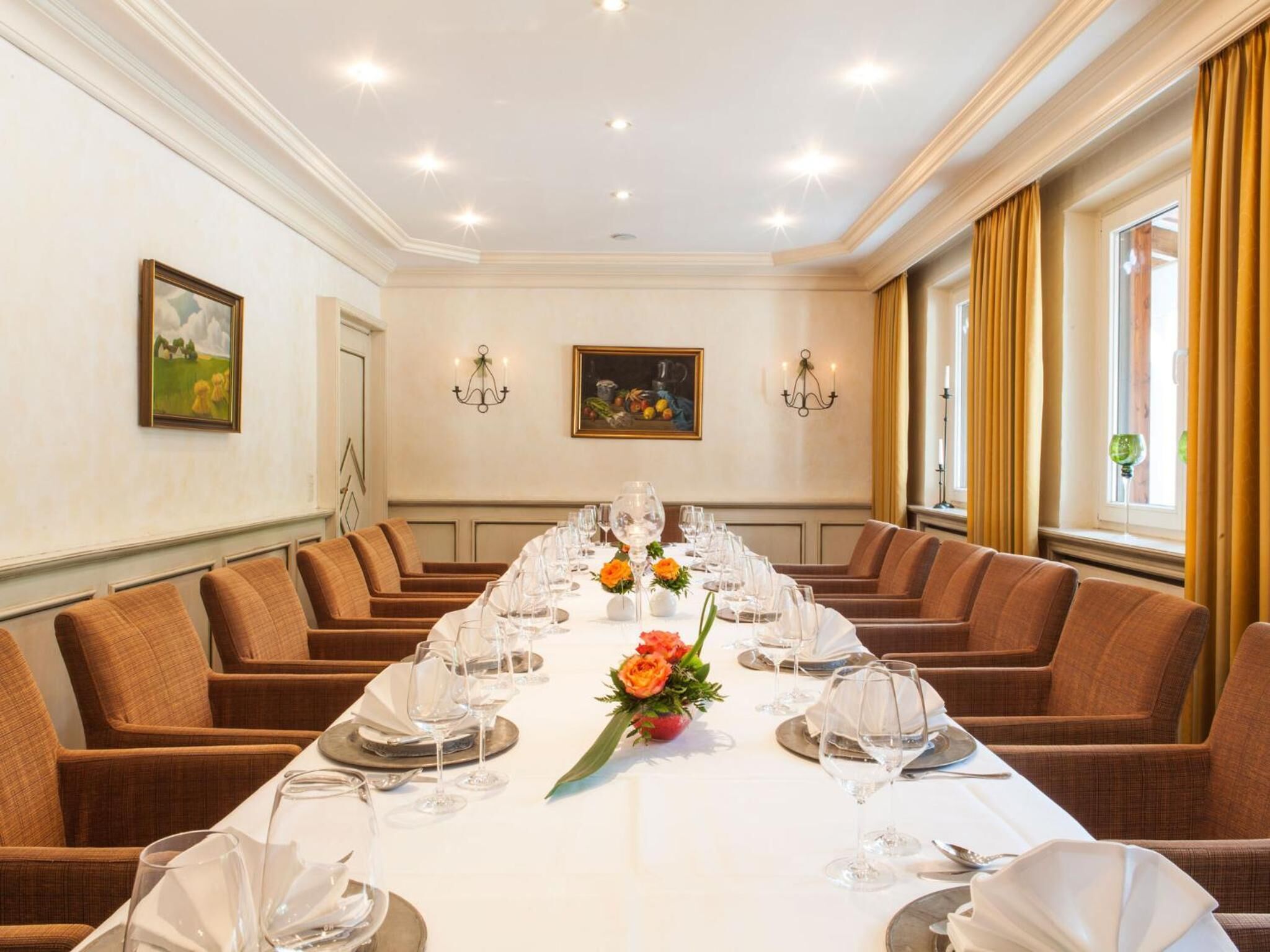 Uma longa mesa de jantar posta para uma refeição formal, com cadeiras estofadas nas laterais, obras de arte nas paredes e um candelabro acima.