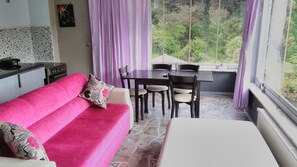 Uma sala de estar aconchegante com um sofá rosa, uma mesa de jantar com cadeiras e vista para o verde através de grandes janelas.