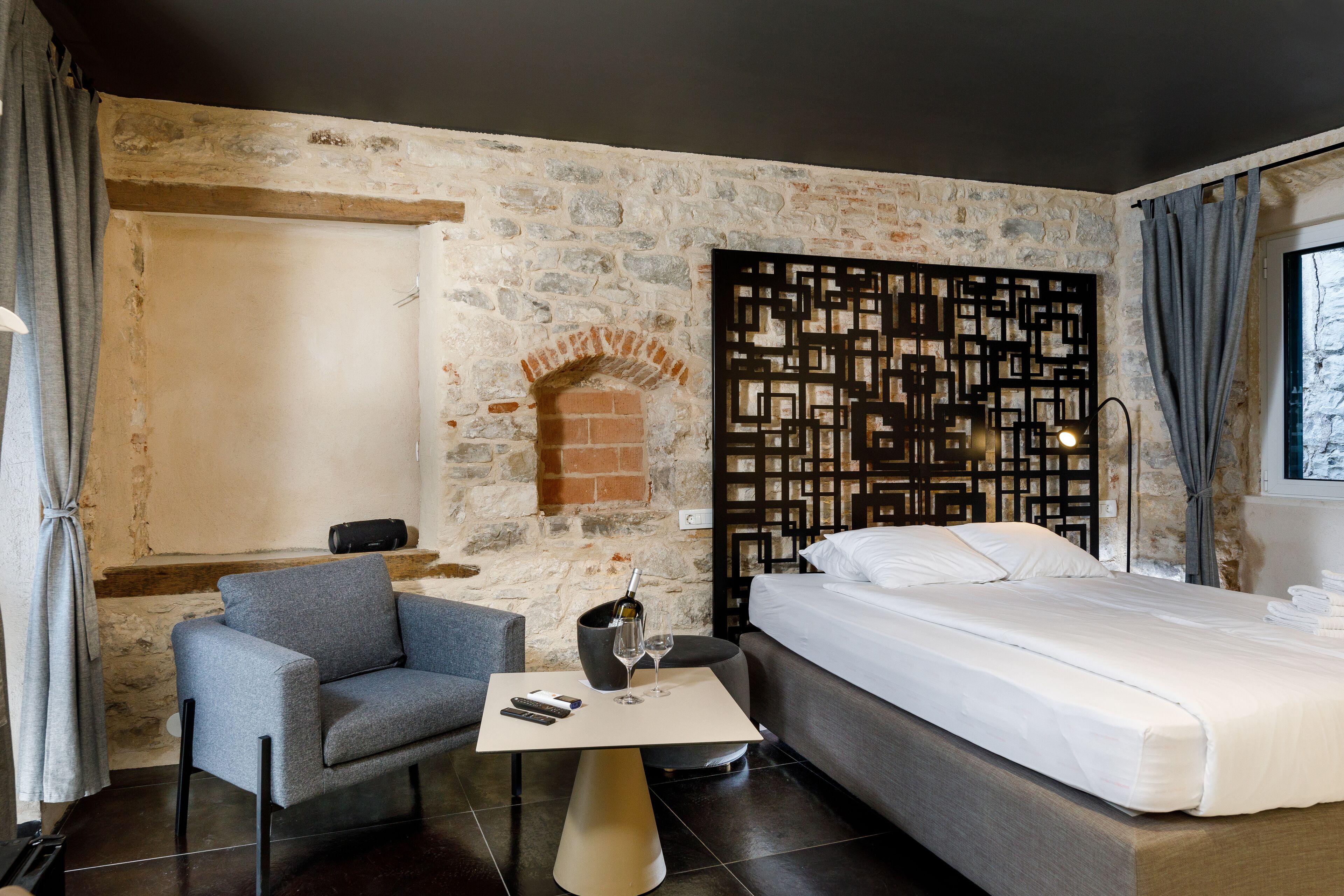 Quarto com parede de pedra, lareira de tijolos, cama, poltrona cinza, mesa pequena com uma garrafa, copos e controle remoto.