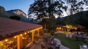 Uma área de pátio ao ar livre com mesas e cadeiras, rodeada por plantas em vasos e árvores.