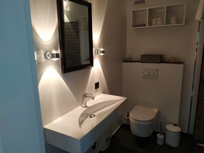 Casa de banho moderna com lavatório branco, espelho, sanita e prateleira com produtos de higiene pessoal.
