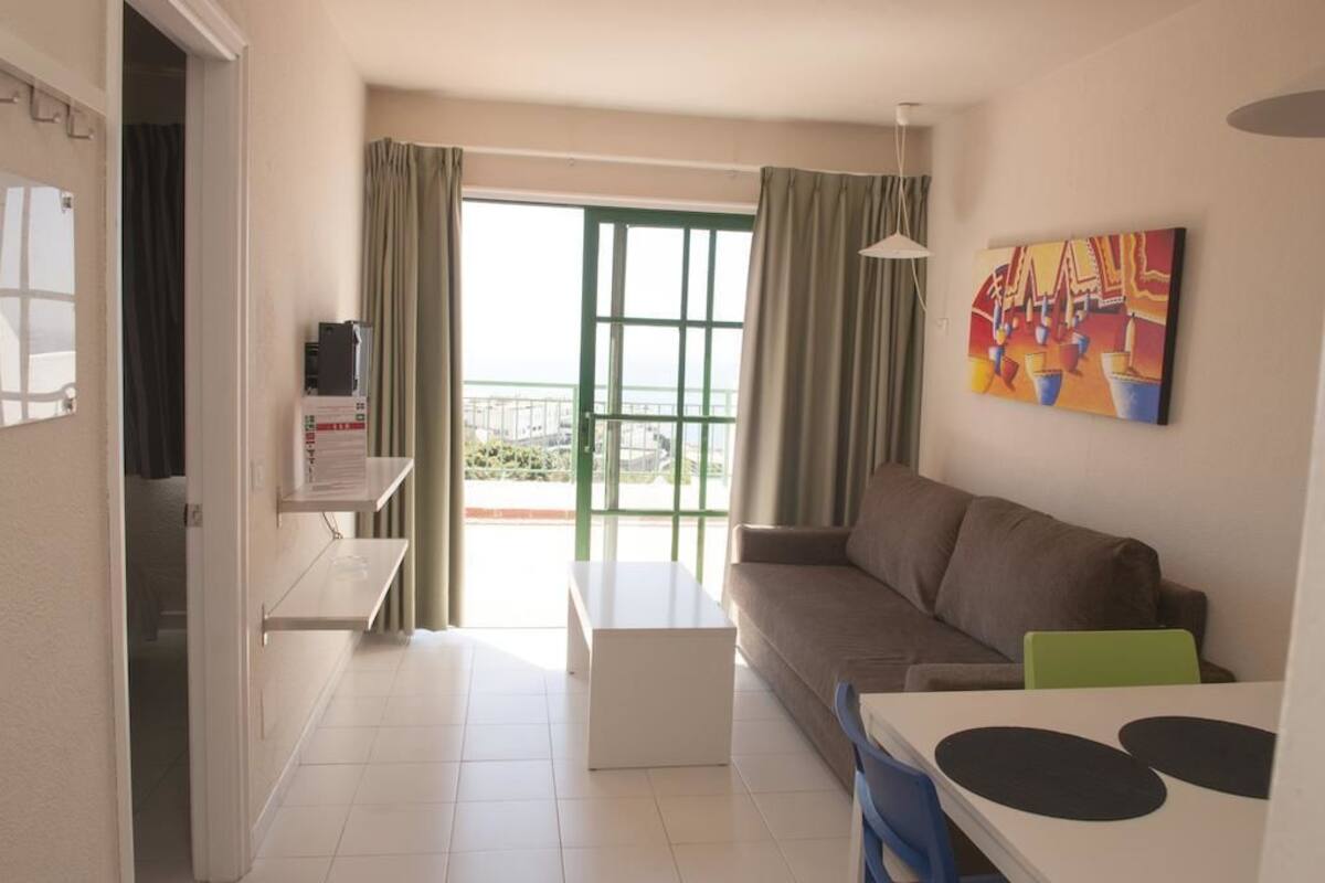 Uma sala de estar com sofá, uma mesa de centro, um quadro na parede e vista através de uma janela grande.