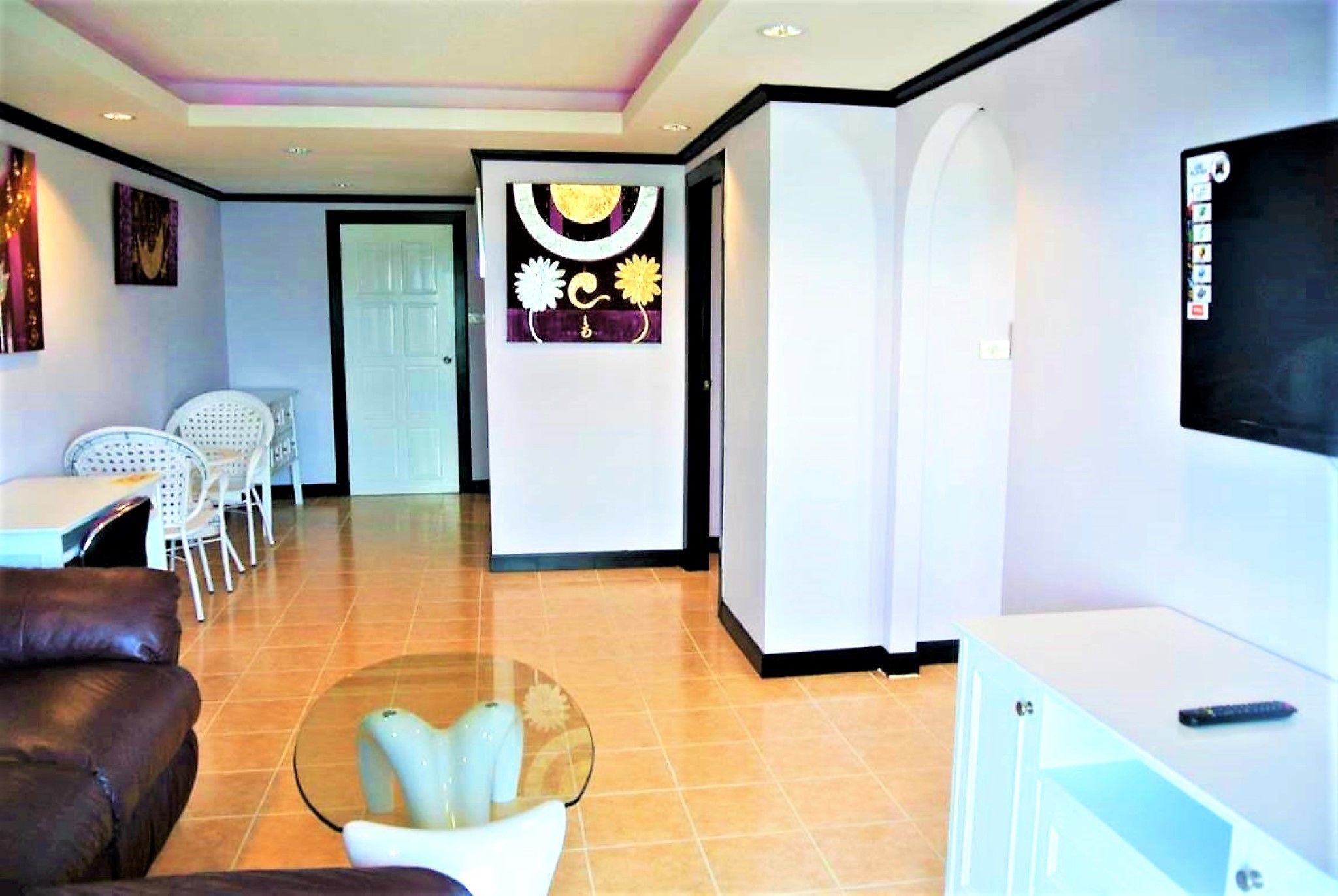 Uma sala de estar moderna com um sofá de couro marrom, uma mesa de centro de vidro e uma TV de tela plana. Há uma área de jantar com cadeiras e mesa brancas.