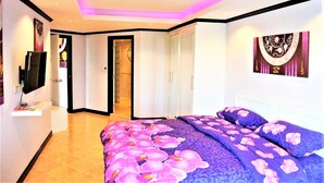 Um quarto com cama, coberta com espalha floral em tons de roxo e rosa, televisão de tela plana em um suporte, e uma porta que dá para um banheiro.