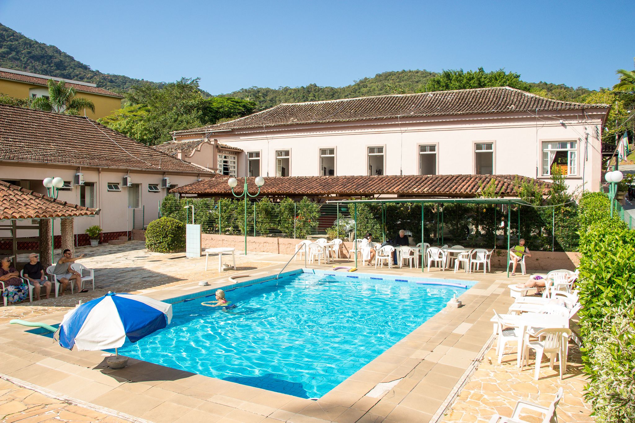 Uma piscina com um guarda-sol azul e branco, rodeada por cadeiras e mesas brancas, com um edifício ao fundo.