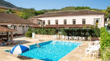 Uma piscina com um guarda-sol azul e branco, rodeada por cadeiras e mesas brancas, com um edifício ao fundo.