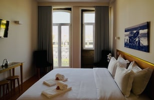 Quarto de hotel com cama, duas cadeiras, escrivaninha e uma janela grande com cortinas.