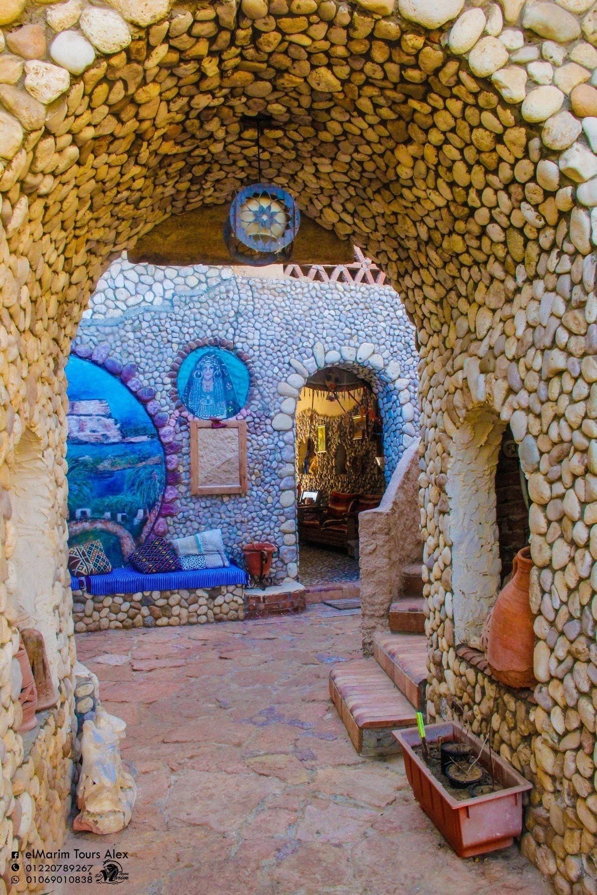 Uma estrutura de pedra arqueada com um mural em mosaico, um banco e uma pequena mesa.