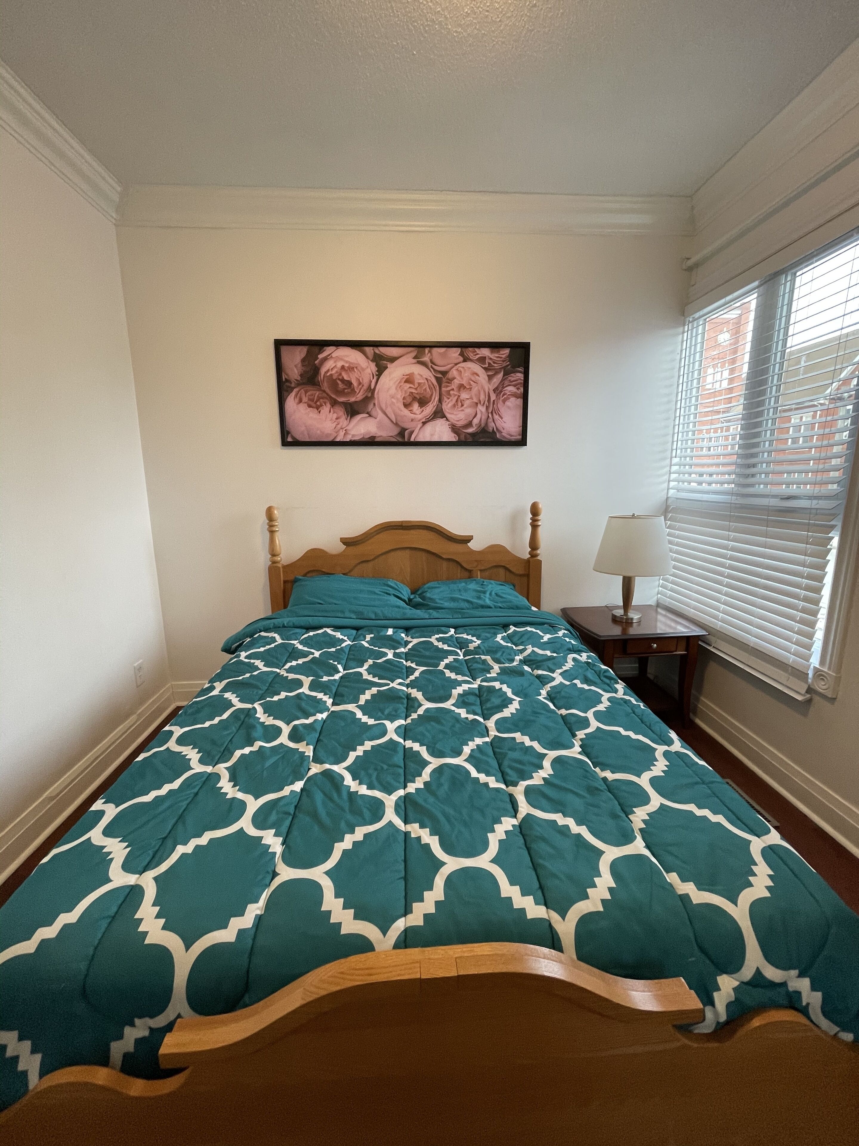 Um quarto com cama, mesa de cabeceira, luminária, janela com persianas e um quadro com flores na parede.