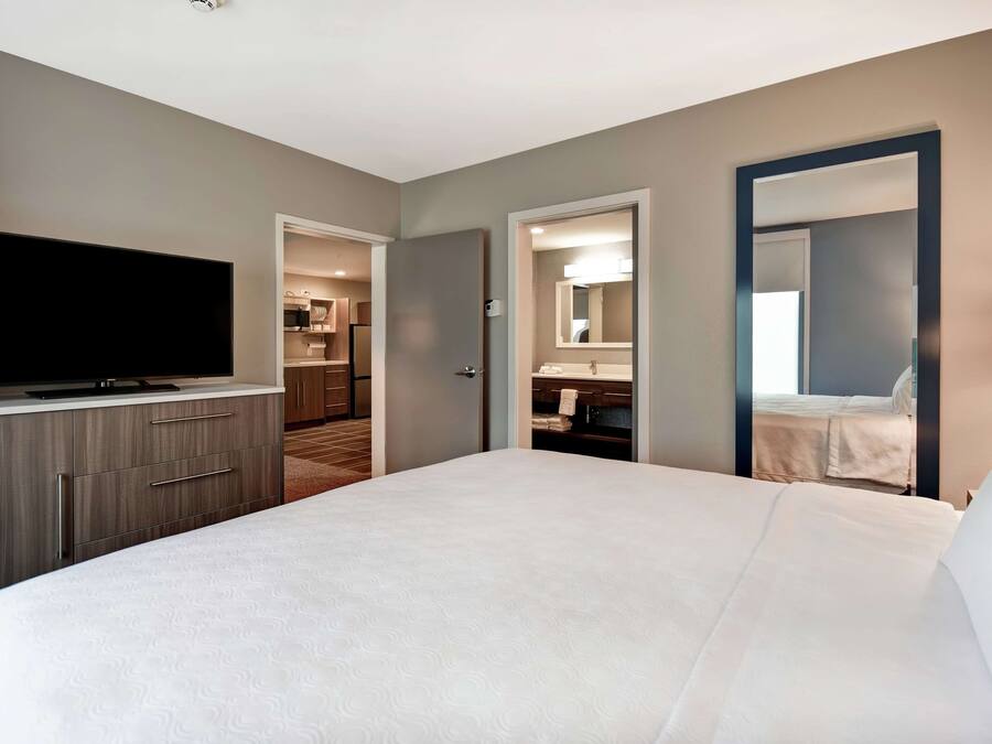 Quarto de hotel com uma cama grande, televisão em uma cômoda, banheiro com espelho e uma kitchenette ao fundo.