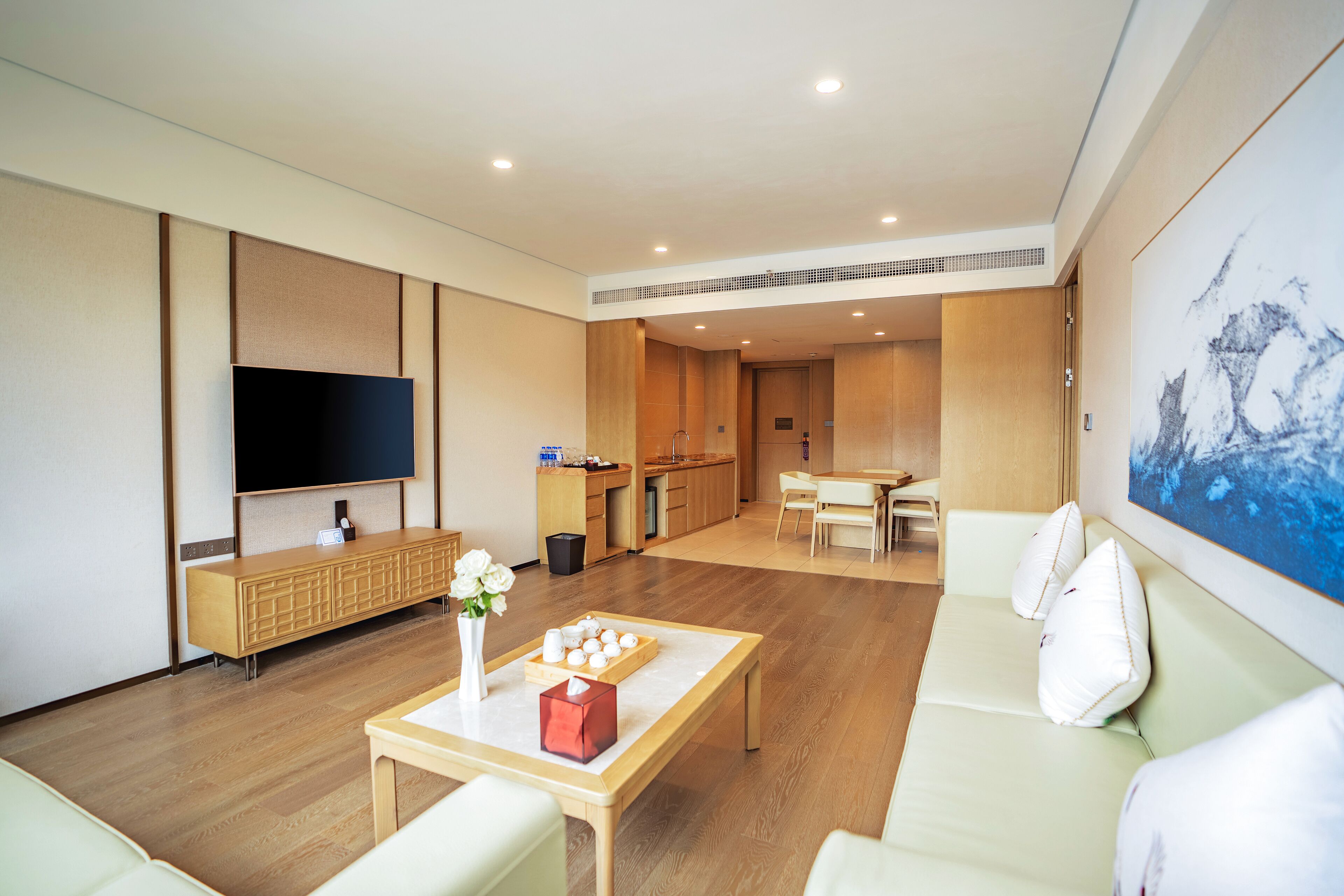 Quarto de hotel moderno com televisão de tela plana, mobiliário de madeira, área de refeições e um quadro na parede.