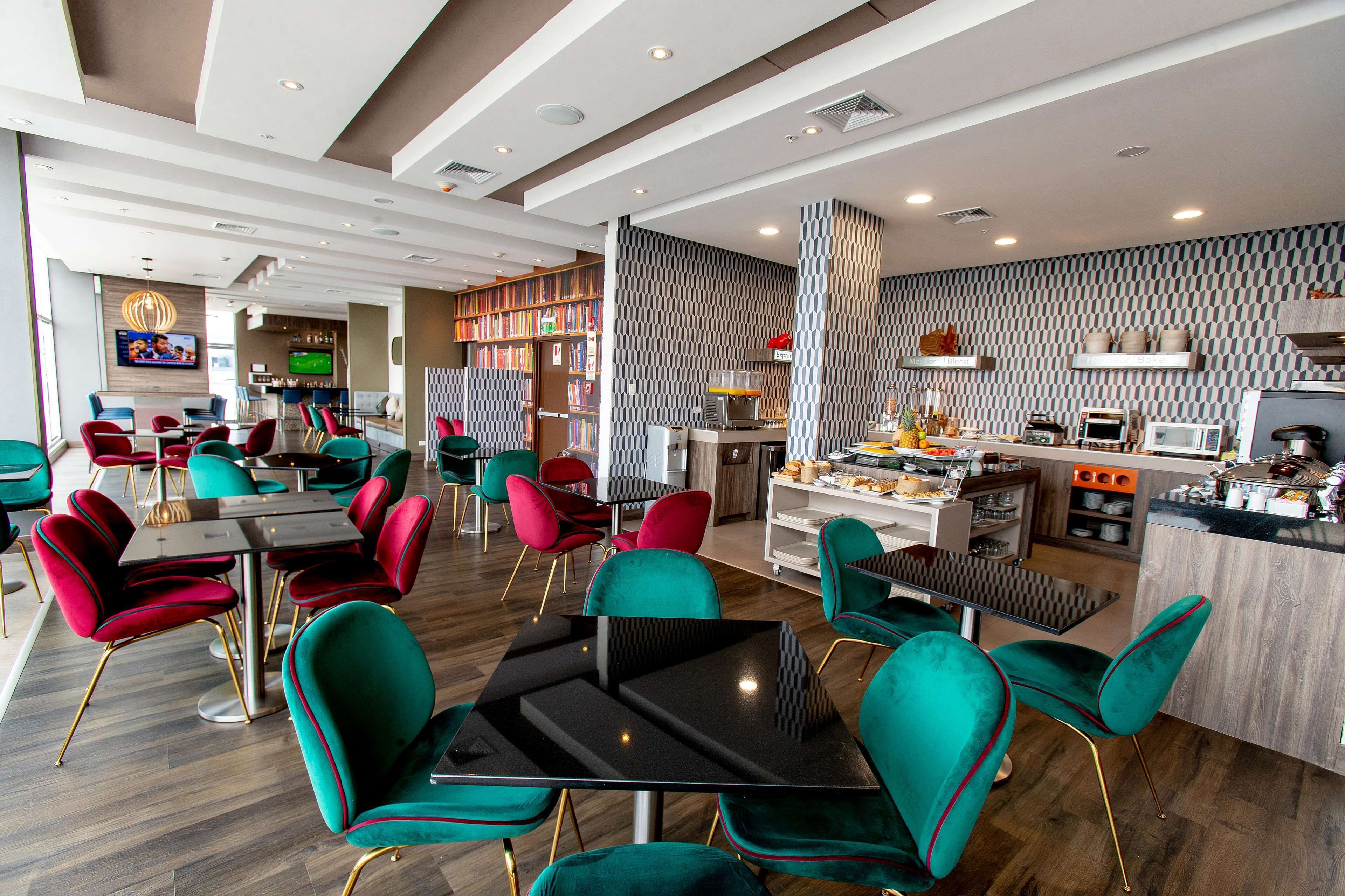 Um restaurante moderno com cadeiras verde-água e vermelhas, uma área de bar e cozinha visível ao fundo.