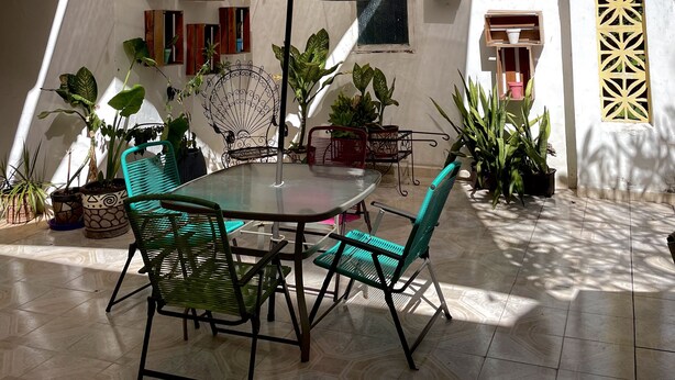 Um pátio com mesa e cadeiras, rodeado por plantas em vasos e um leque metálico decorativo.