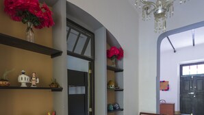 Um corredor com chão xadrez, prateleiras com objetos decorativos, um candelabro e uma porta que dá para outro cômodo.