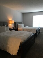 Quarto de hotel com duas camas, uma mesa de cabeceira com abajur e uma janela grande.