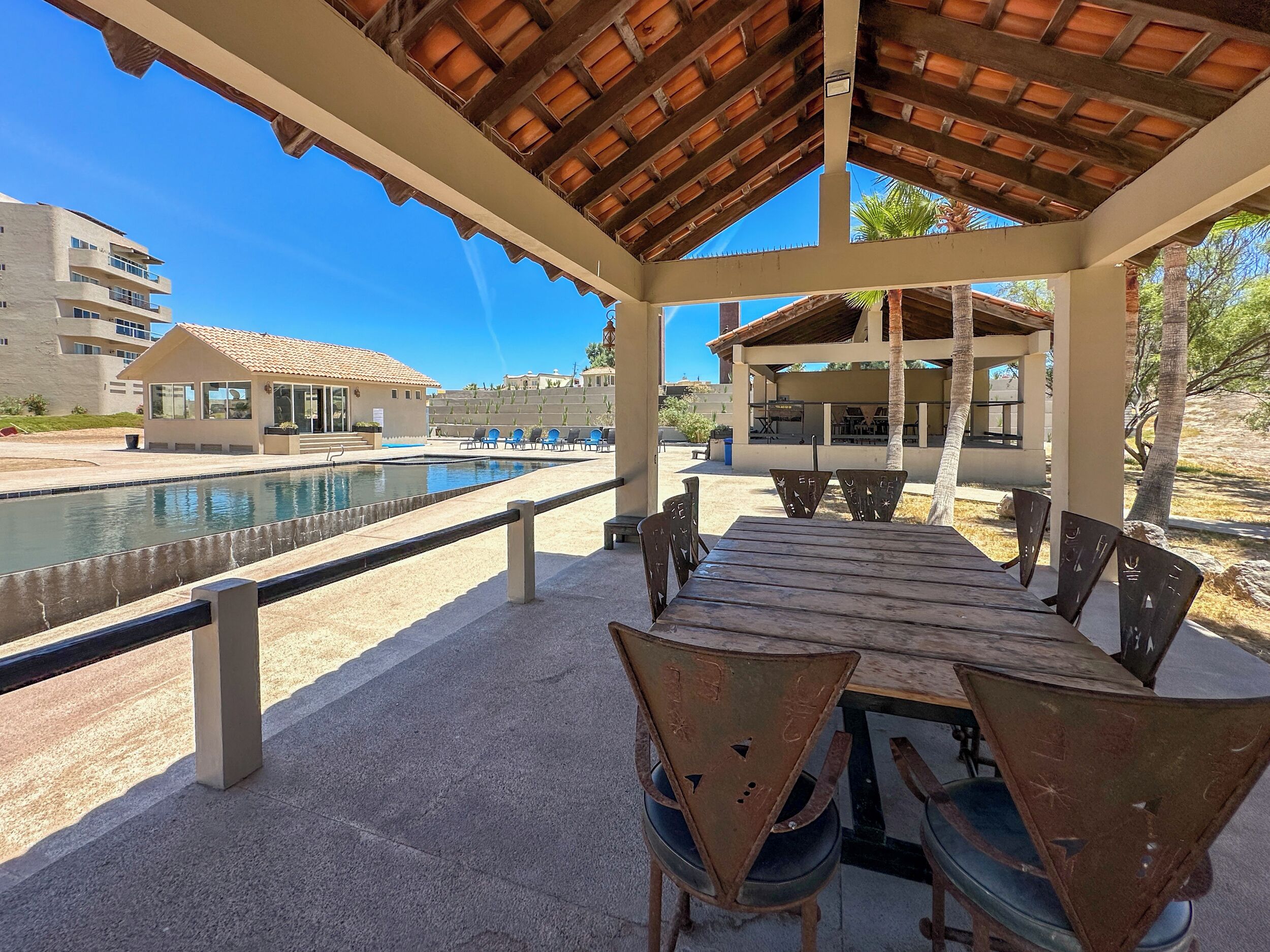 Uma área externa coberta com piscina, mesa e cadeiras, e vista para um edifício ao fundo.