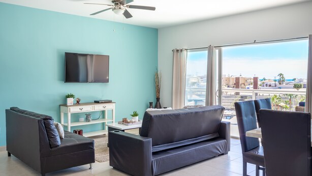 Uma sala de estar com parede verde-água, um ventilador de teto, uma TV de tela plana, um sofá, uma mesa de centro e vista da cidade por grandes janelas.