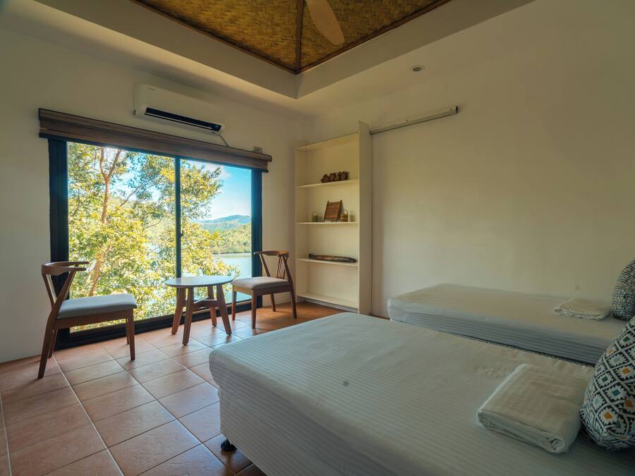 Quarto com uma janela grande, uma cama, uma cadeira, uma mesa pequena e uma estante com objetos decorativos.