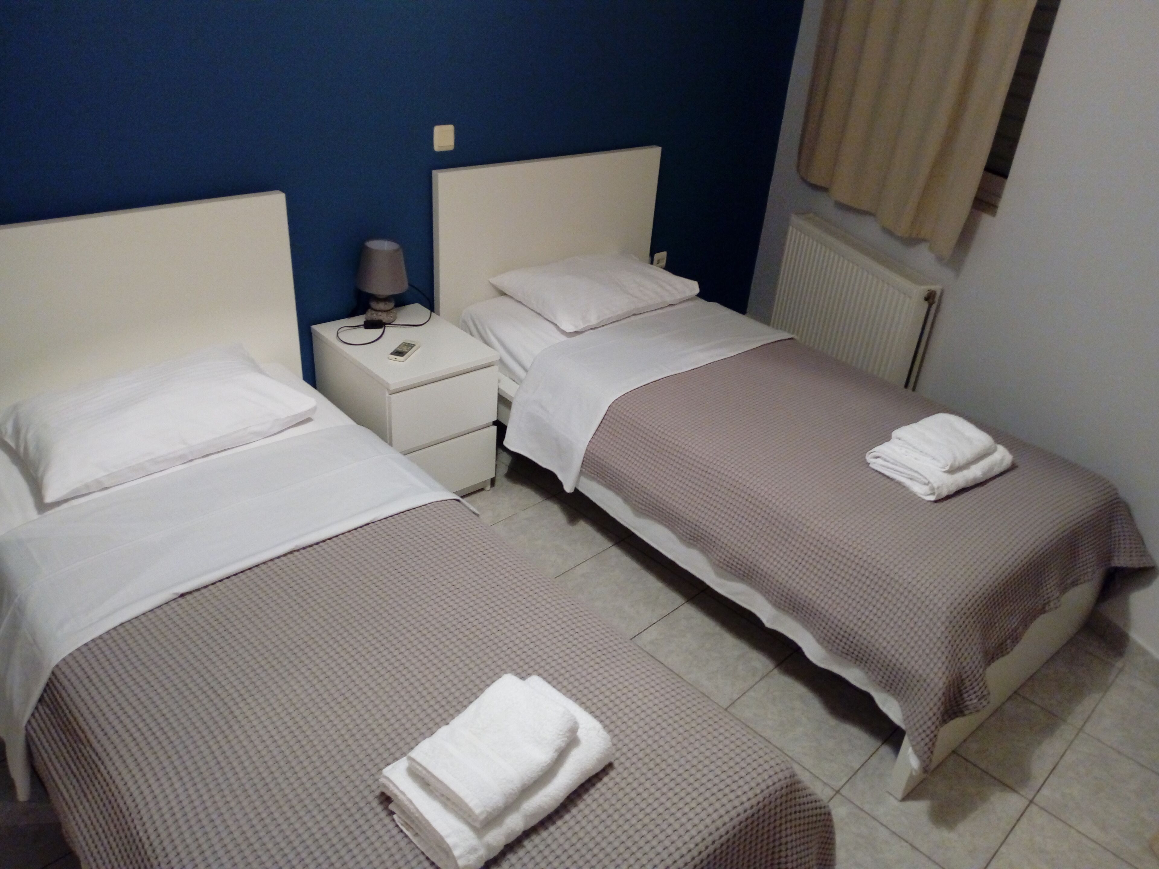 Quarto de hotel com duas camas de solteiro, uma mesa de cabeceira com abajur e uma janela com cortinas.
