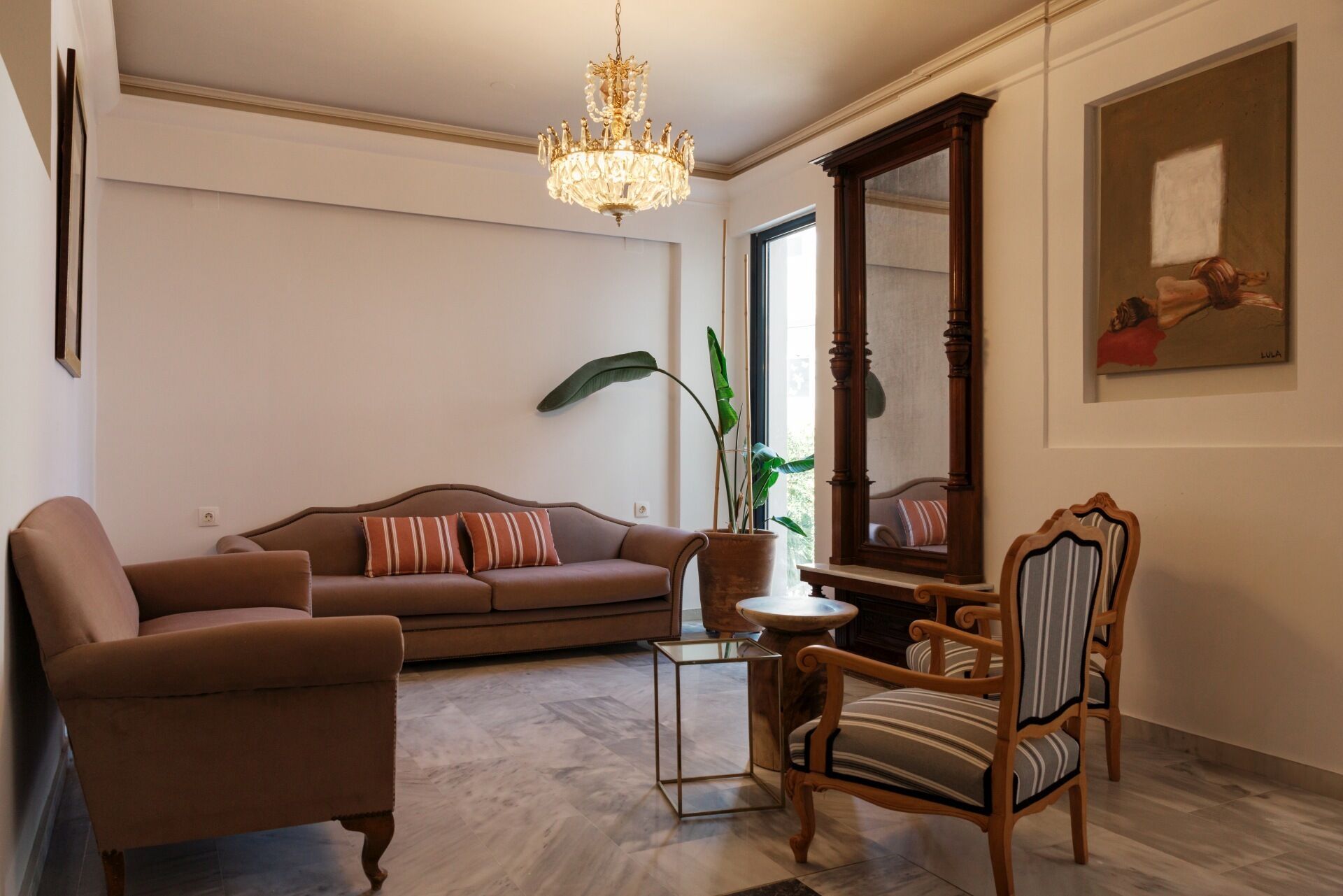 Uma sala de estar com um sofá marrom, uma cadeira de madeira, uma mesa de centro de vidro, um espelho grande e um candelabro.