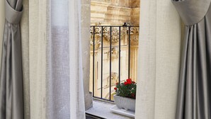 Uma varanda com vista para um balaustrada de pedra ornamentada, um vaso com plantas e flores vermelhas, e um chão de azulejos.