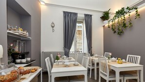 Uma área de refeições com mesas e cadeiras brancas, um buffet com doces e uma vista da cidade através de grandes janelas.