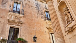 Fachada de um edifício histórico com colunas ornamentadas, uma varanda com grades de ferro forjado e uma estátua.