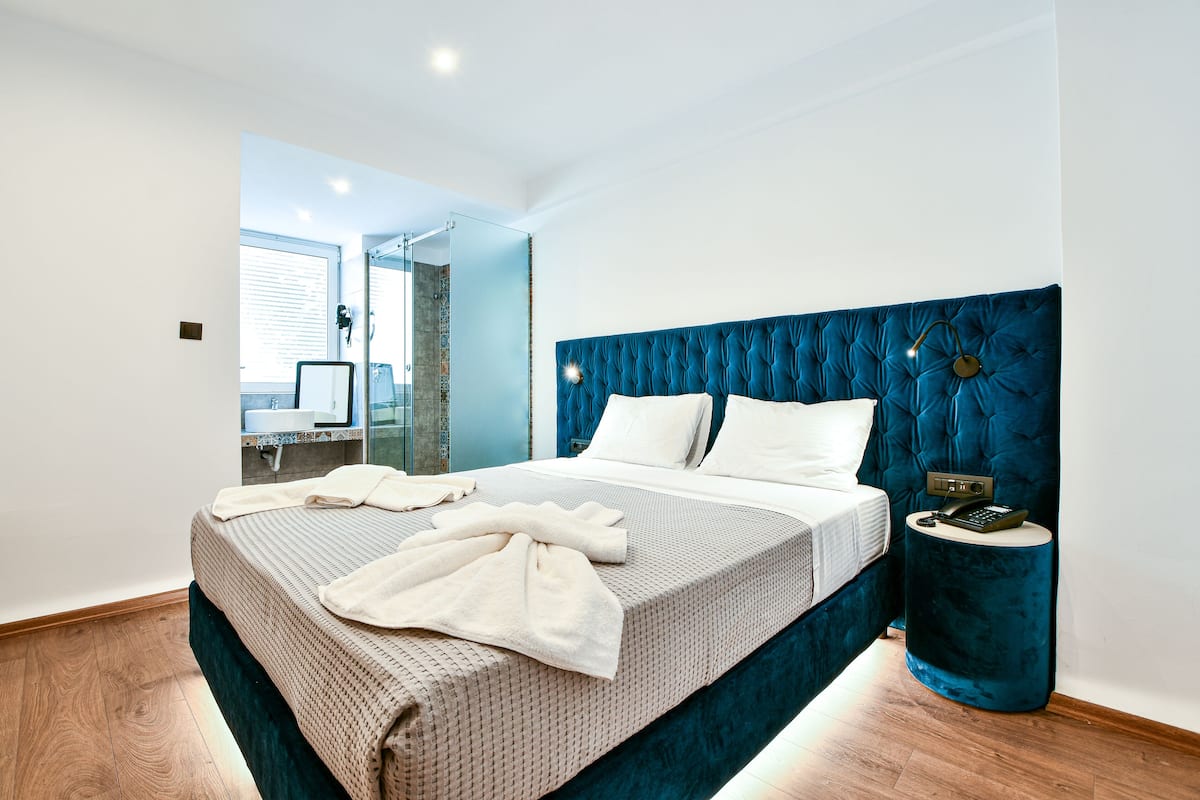 Quarto moderno com uma cama grande, cabeceira azul, mesa de cabeceira com um telefone e área de chuveiro em vidro.