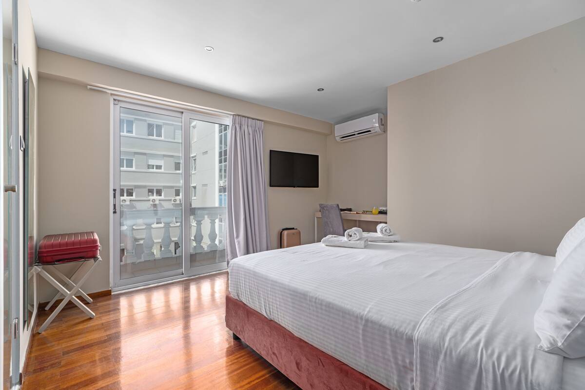 Quarto de hotel com uma cama grande, televisão de tela plana, ar condicionado, uma mesinha com luminária e porta de vidro corrediça que dá para uma varanda externa.