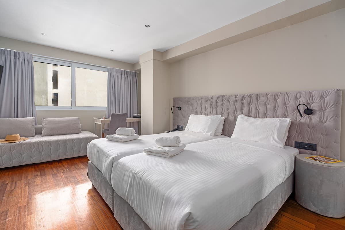 Quarto de hotel com duas camas, um sofá, uma mesa de cabeceira, uma luminária e uma janela com cortinas.