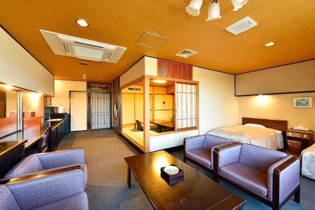Quarto de hotel com uma cama, duas poltronas, uma mesinha, um espelho, uma cozinha compacta e um guarda-roupa com portas de correr.