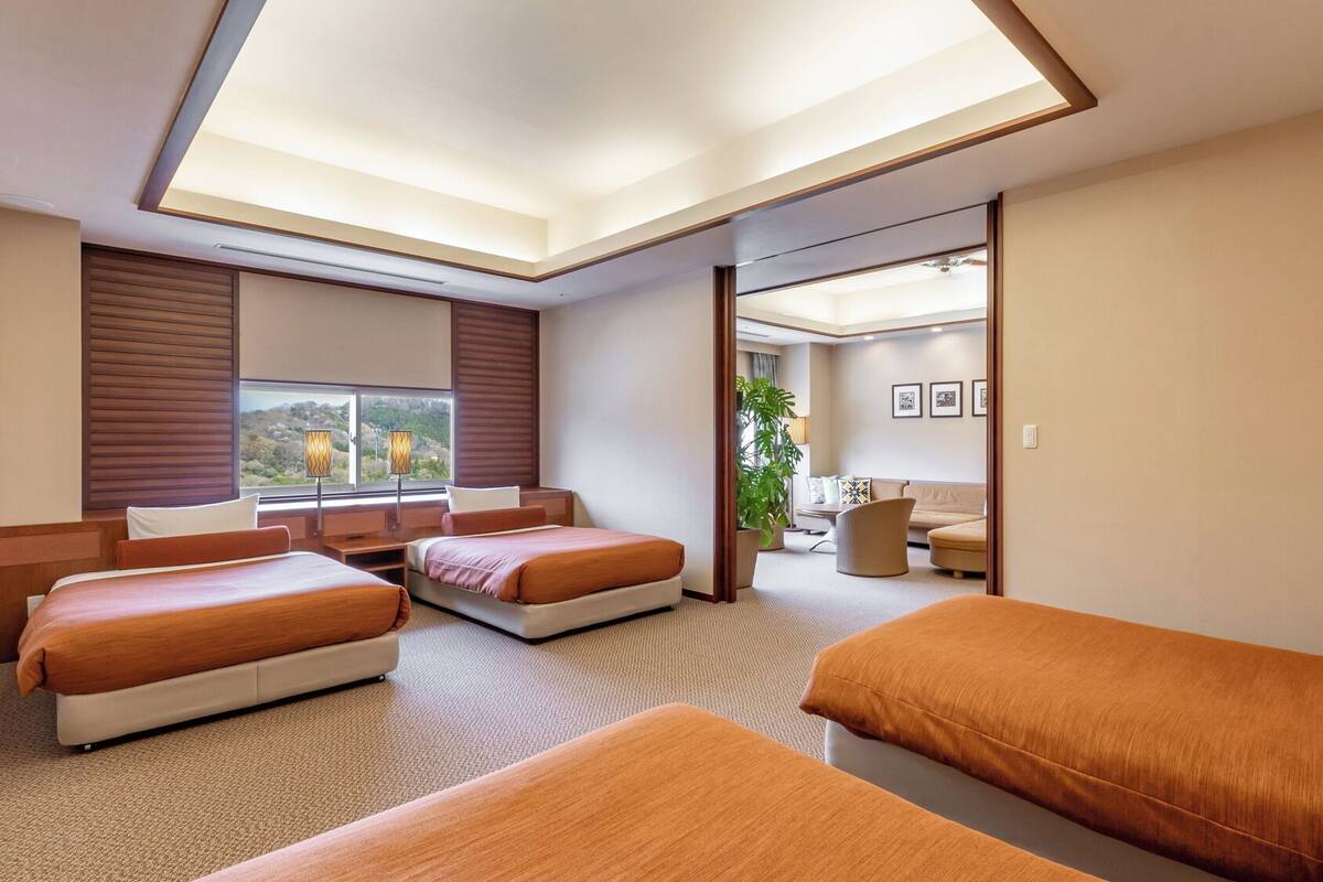 Quarto de hotel com uma cama grande, uma cama menor, um sofá, uma escrivaninha e uma planta.