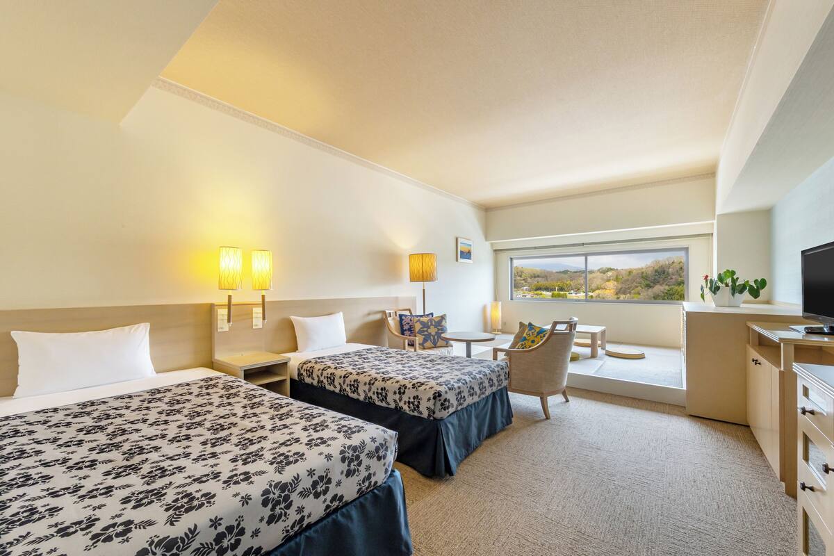 Quarto de hotel com duas camas, uma escrivaninha, uma cadeira, uma televisão e janela com vista.