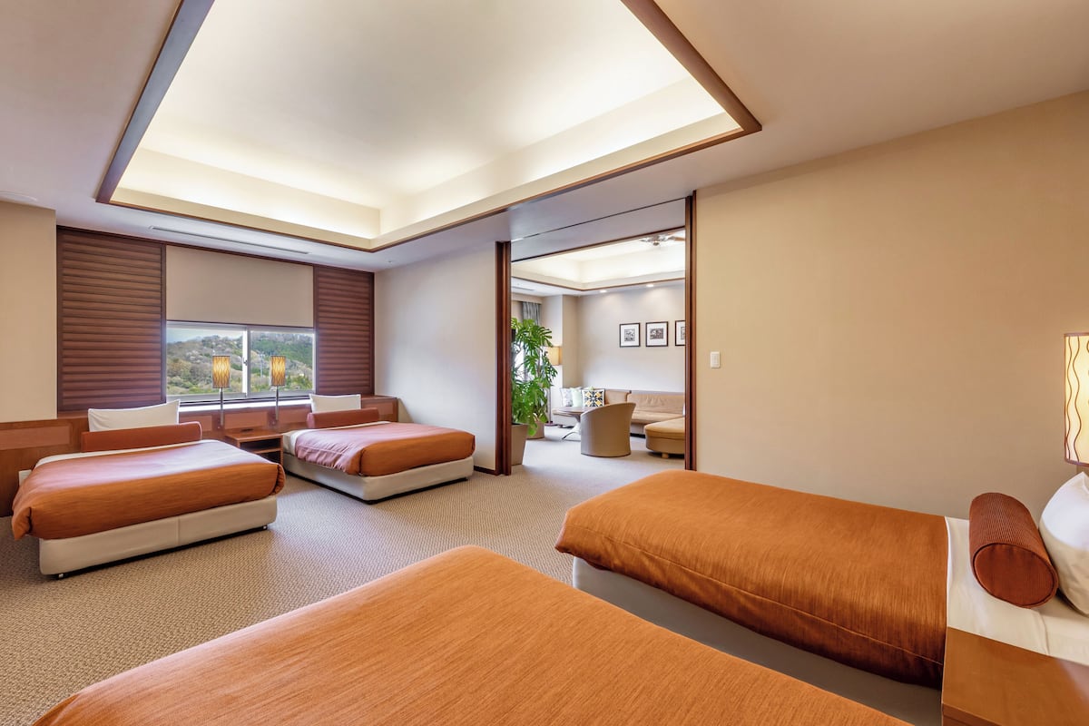 Quarto de hotel com duas camas, um sofá, uma escrivaninha e vista para o exterior.