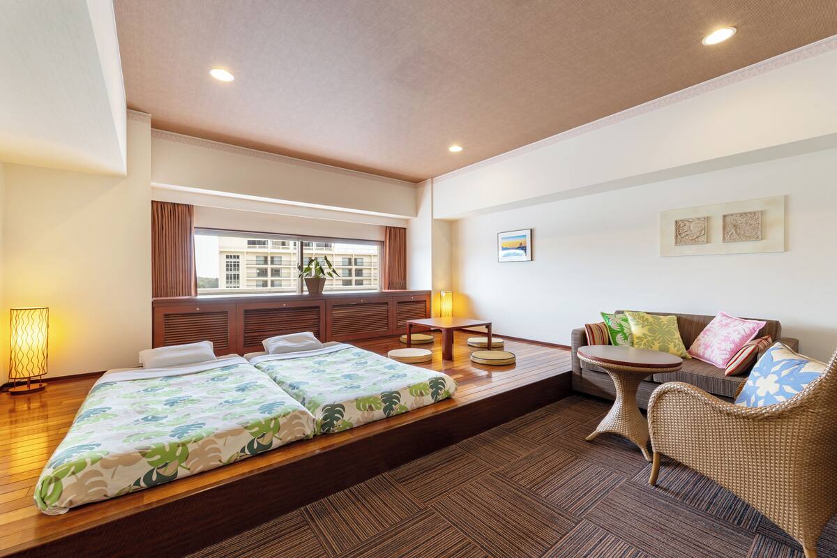 Quarto de hotel com cama, sofá, uma pequena mesa e uma cadeira. Há uma janela com vista para edifícios.