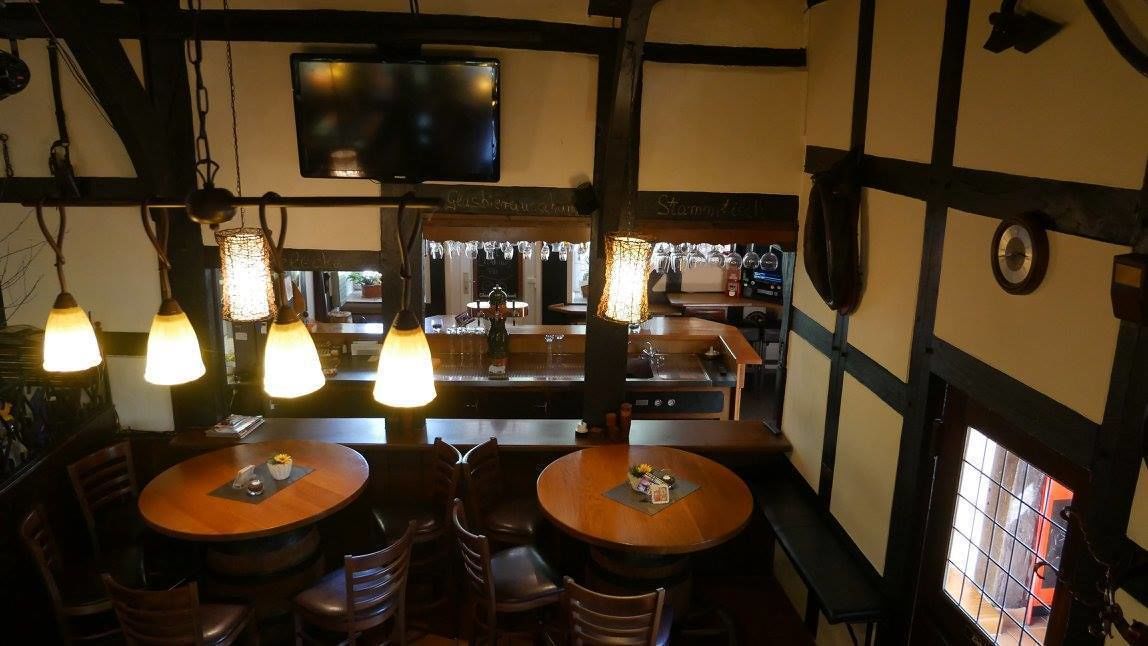 Restaurante com mesas e cadeiras de madeira, luminárias pendentes e uma televisão na parede.