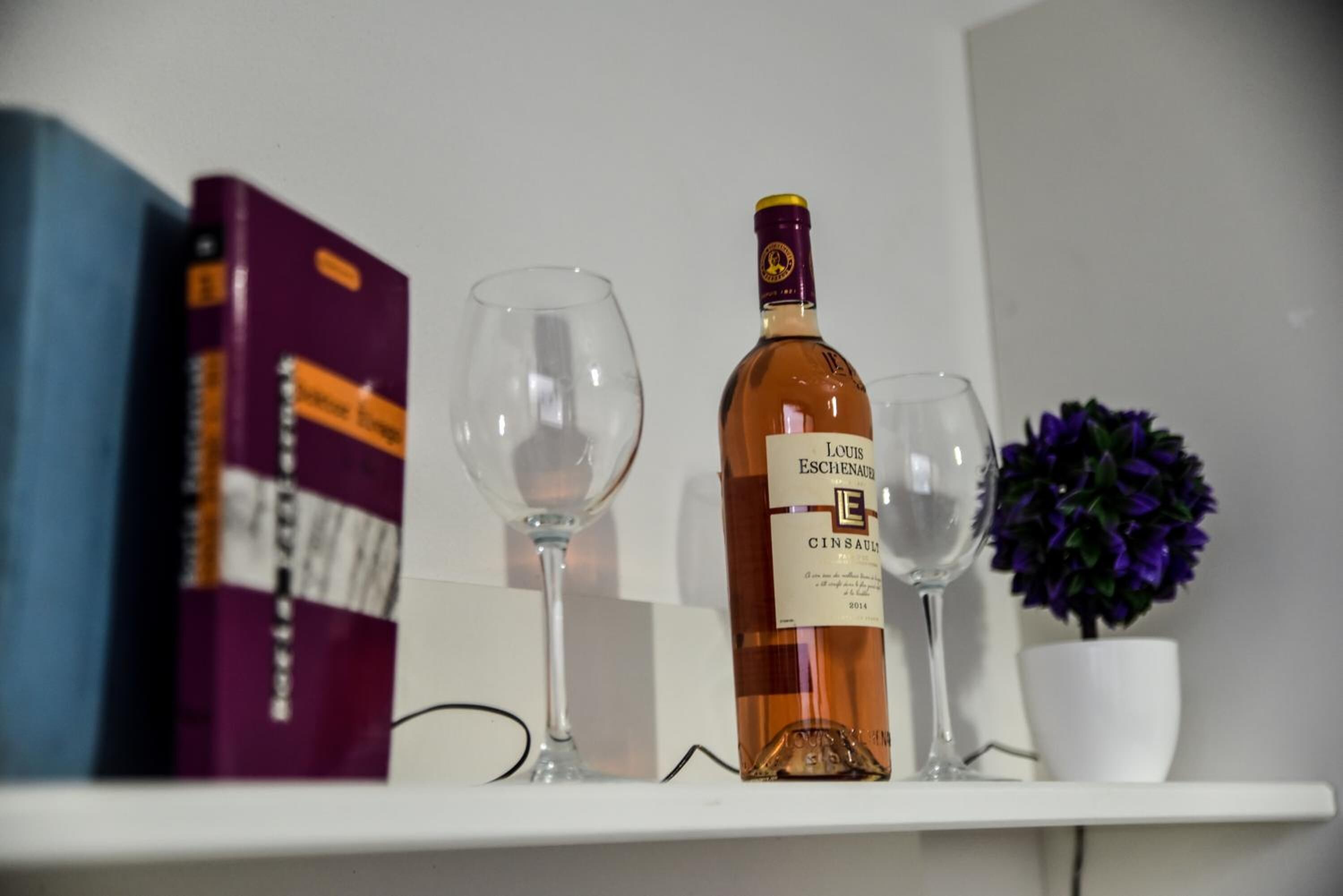 Uma garrafa de vinho Louis Eschener Cinsault 2014 numa prateleira com dois copos de vinho vazios e um livro roxo.