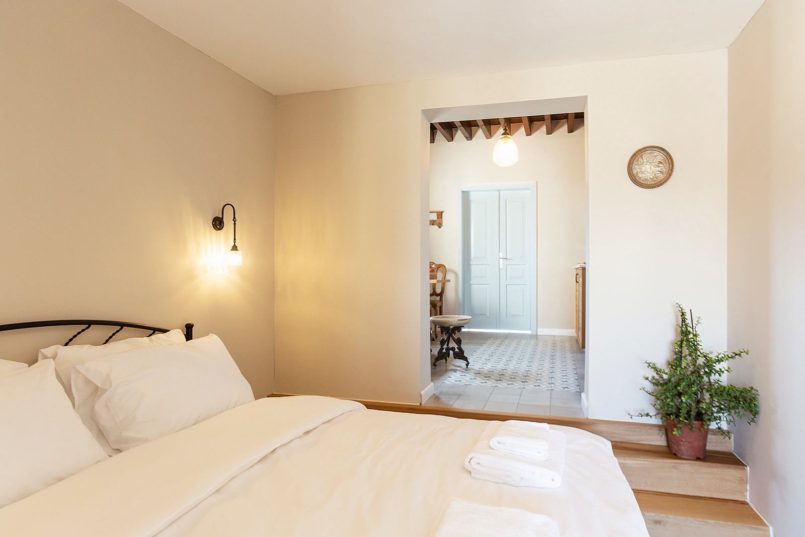 Um quarto com cama, mesa de cabeceira, luminária na parede, planta em vaso e vista para o banheiro por uma porta aberta.