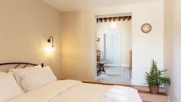 Um quarto com cama, mesa de cabeceira, luminária na parede, planta em vaso e vista para o banheiro por uma porta aberta.
