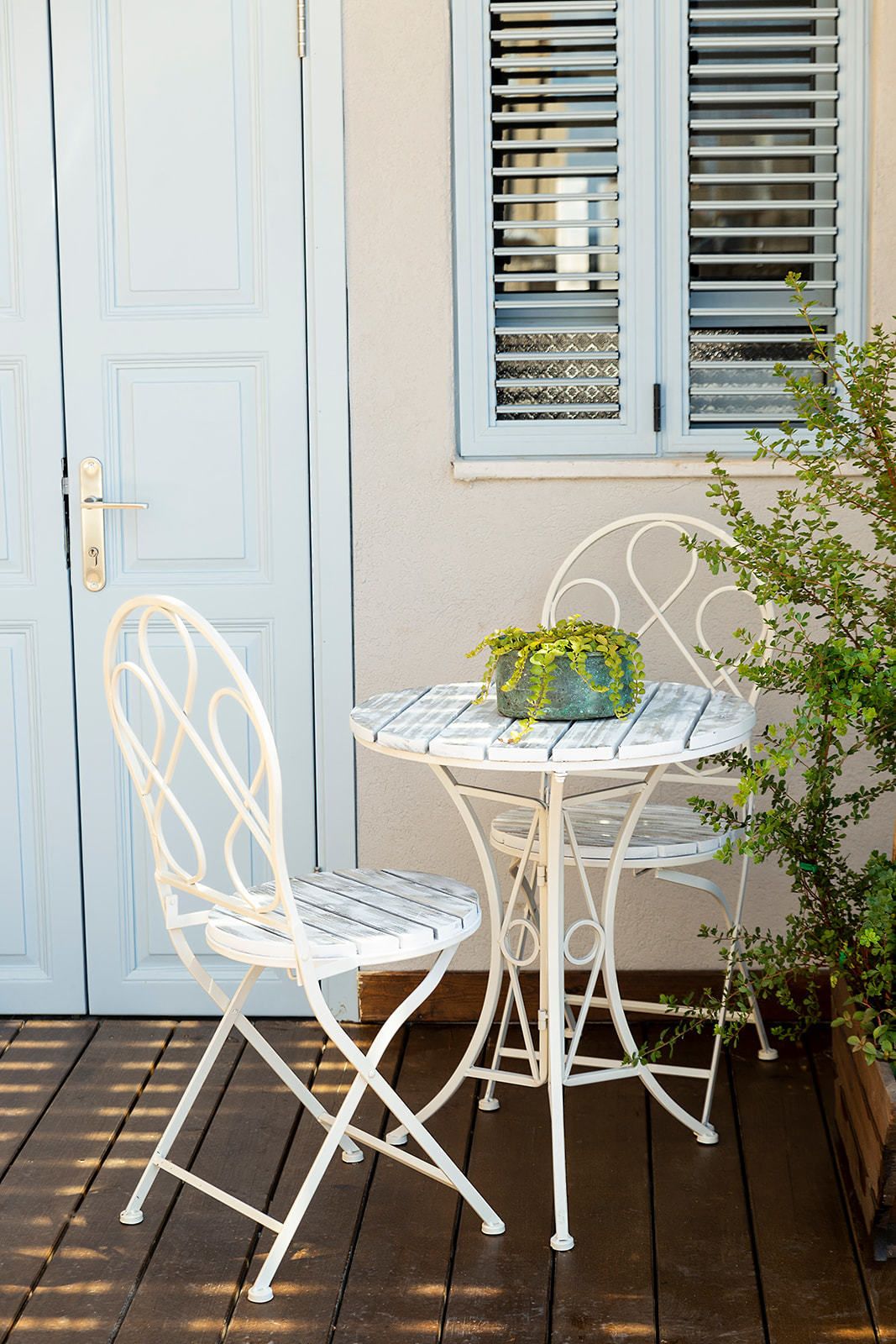 Conjunto de mesa e cadeira de metal branco sobre um deck de madeira, com um vaso de planta ao lado.